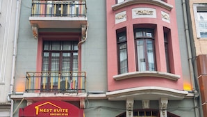 Um edifício de vários andares com uma fachada marcante em vermelho e rosa, uma vitrine visível com manequins e uma placa com 'NEST SUITE' e um número de telefone.