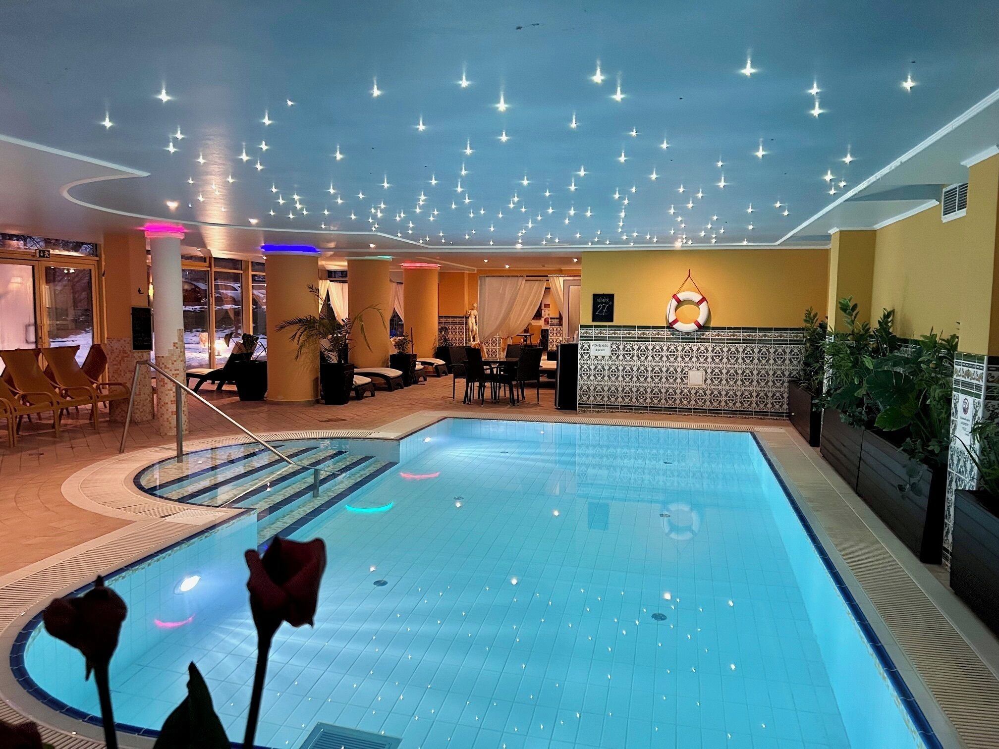 Piscina interior com chão ladrilhado, rodeada por espreguiçadeiras e uma área de estar com mesas.