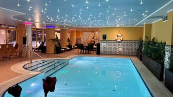 Piscina interior com chão ladrilhado, rodeada por espreguiçadeiras e uma área de estar com mesas.