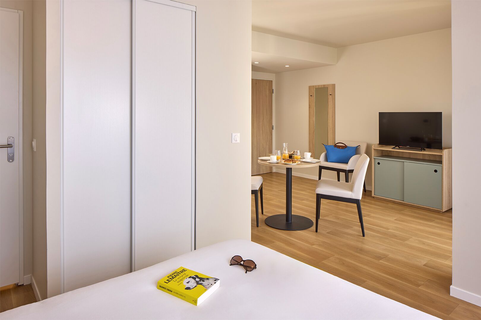 Quarto de hotel moderno com cama, área de jantar com mesa e cadeiras, televisão e guarda-roupa.