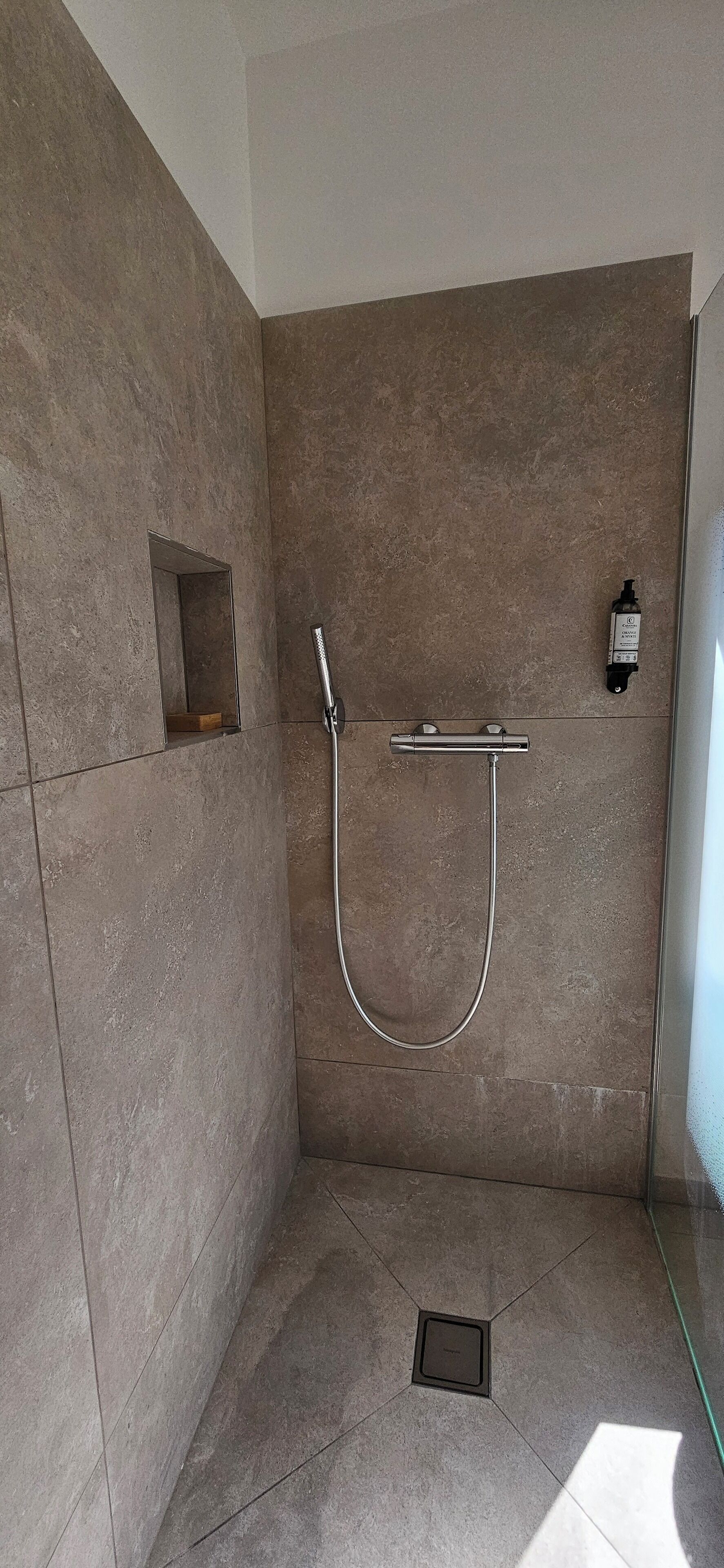 Um chuveiro moderno com um chuveiro manual, um pequeno prateleira e uma garrafa fixada na parede.