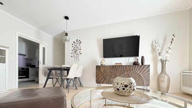 Uma sala de estar moderna com uma mesa de centro de vidro, um sofá, uma TV fixada na parede e uma cozinha ao fundo.