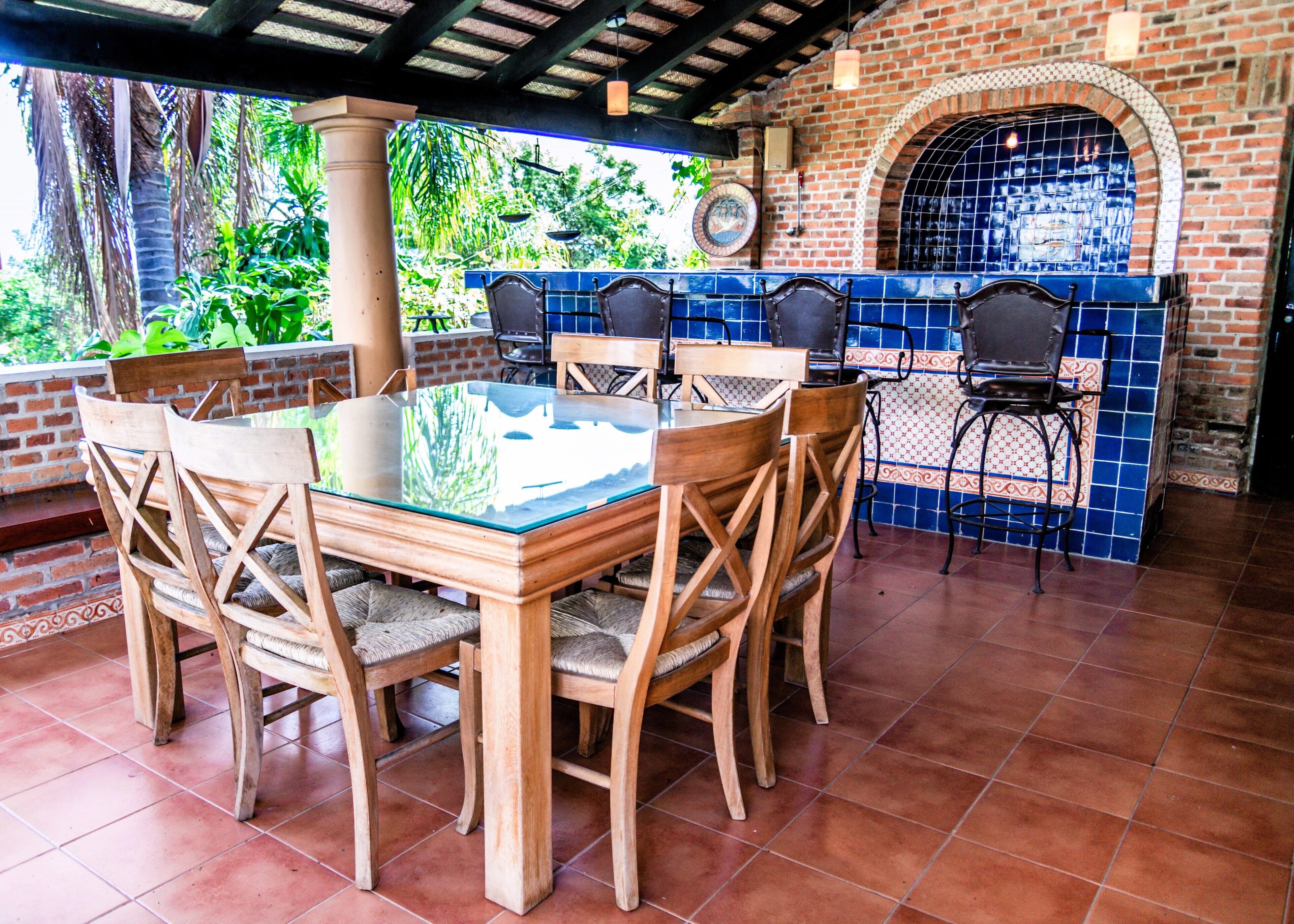 Um pátio externo com mesa de vidro rodeada por cadeiras de madeira, parede de tijolos e revestimento de azulejos.
