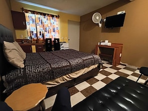 Quarto de hotel com cama, escrivaninha, cadeira, ventilador e televisão.