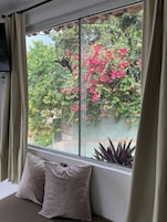 Quarto com uma janela grande, uma cama com duas almofadas e vista para um jardim com flores cor-de-rosa.
