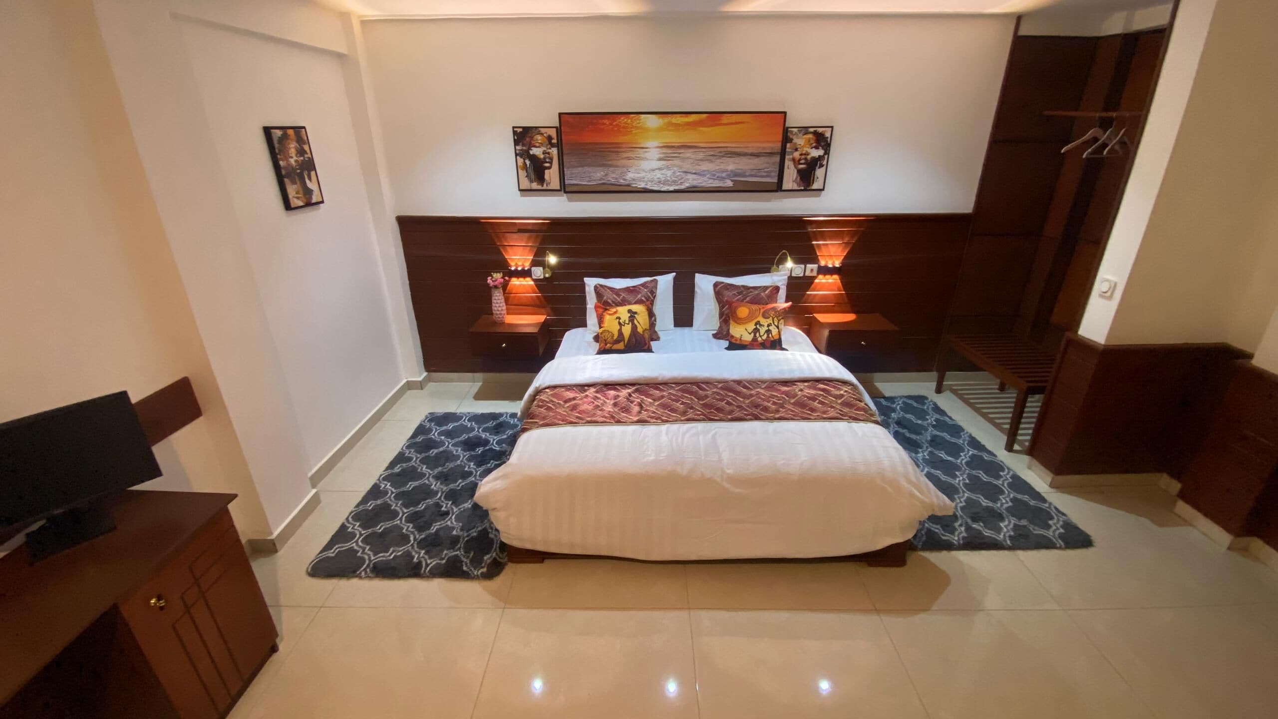 Quarto de hotel com uma cama grande, duas luminárias de cabeceira, um quadro acima da cama e uma cabeceira de madeira.
