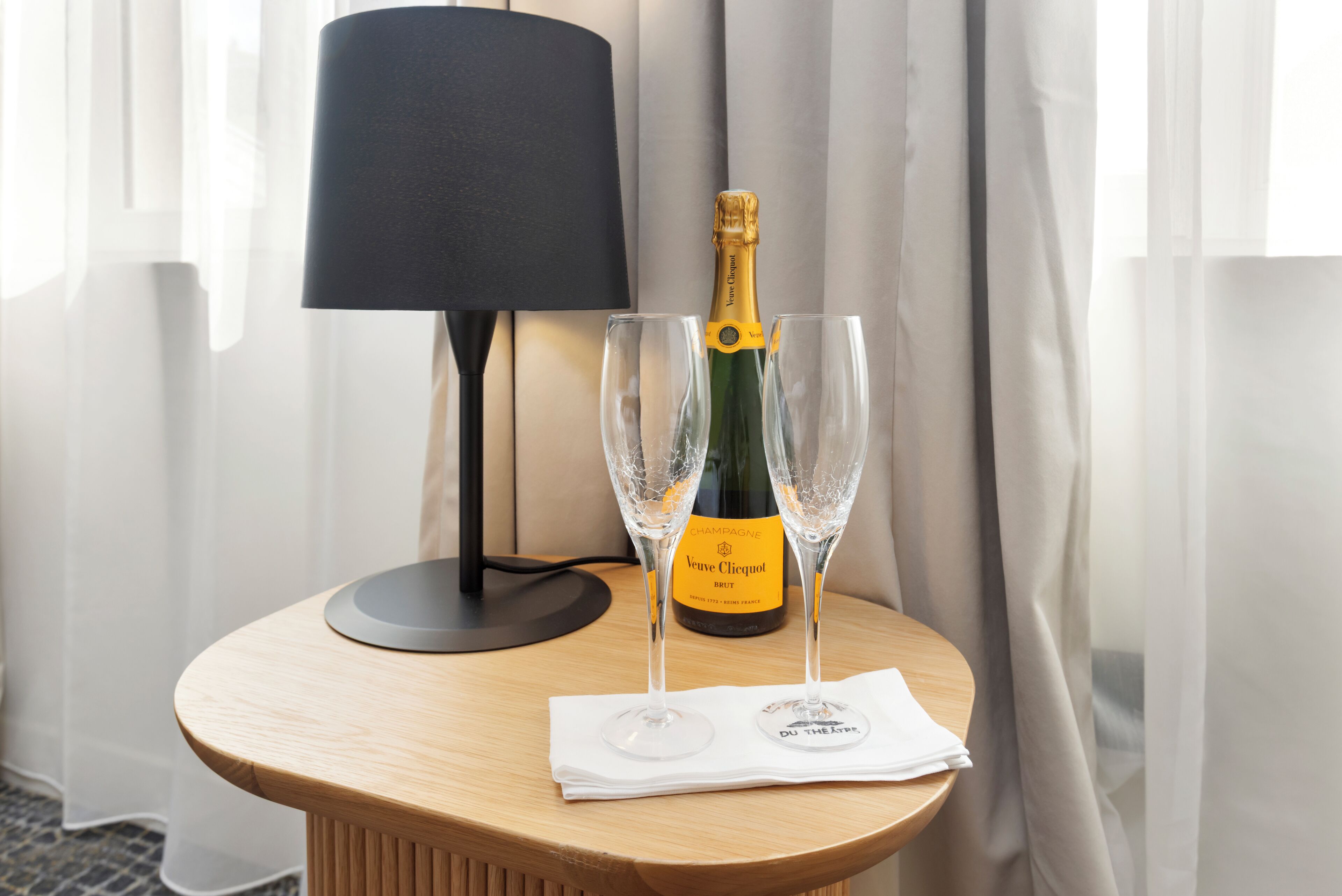 Uma garrafa de champanhe Veuve Clicquot e dois copos sobre uma mesa de madeira com uma luminária preta, em frente a uma janela com cortinas finas.