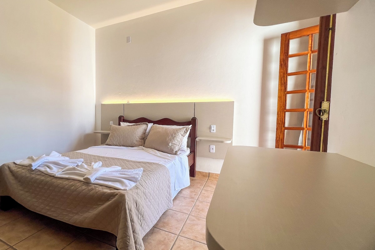 Cama bem arrumada com cabeceira, mesa de cabeceira e porta de madeira com painel de vidro.
