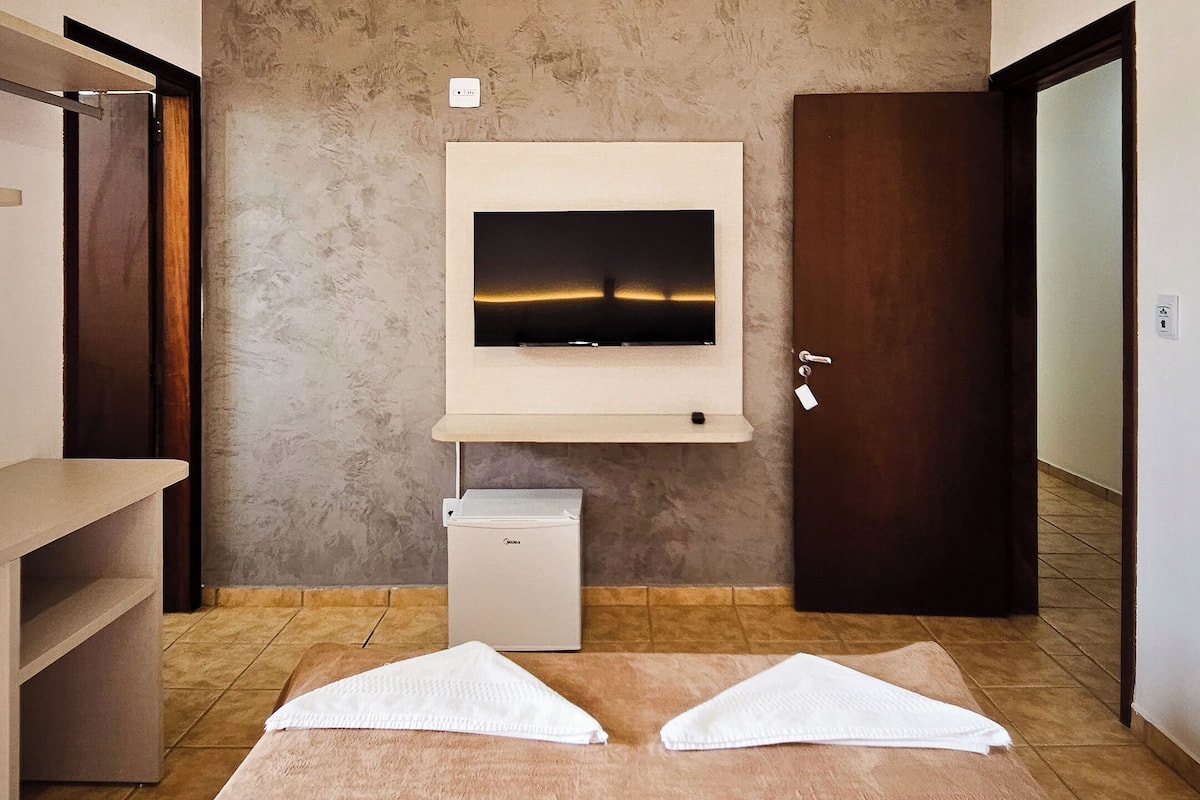 Quarto de hotel com cama, televisão na parede, ventoinha no teto e minibar.