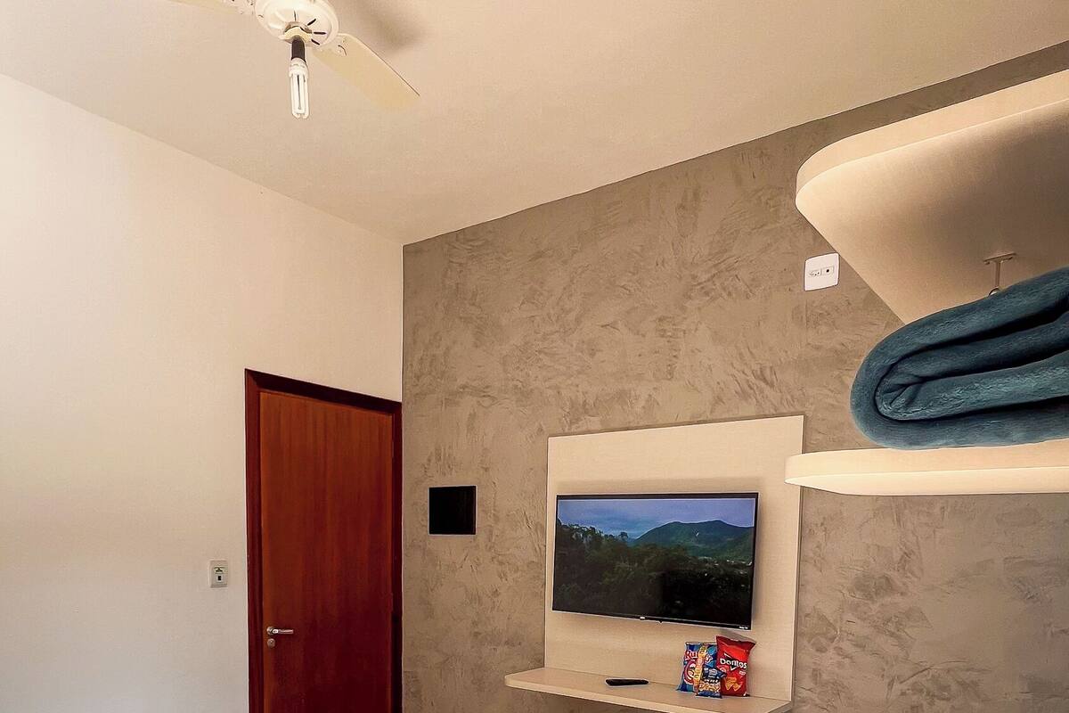 Quarto de hotel com cama, televisão na parede, ventilador de teto e pequena área de cozinha.