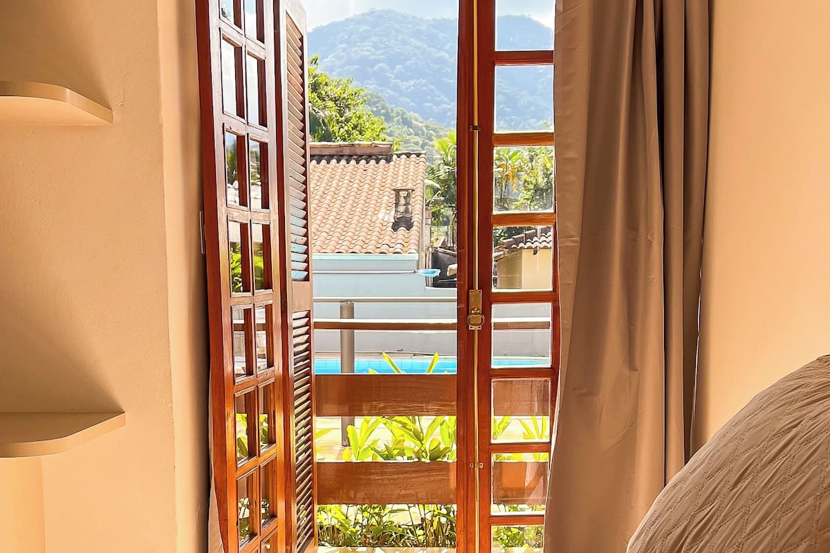 Quarto de hotel com cama, vista para piscina e montanhas, e porta de madeira com cortina.