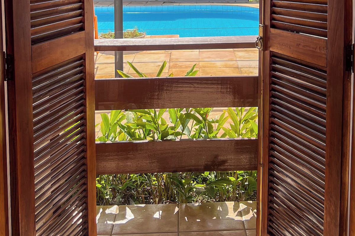 Uma porta de madeira aberta dá para uma varanda com vista para uma piscina, uma montanha e um edifício com telhado de azulejos.