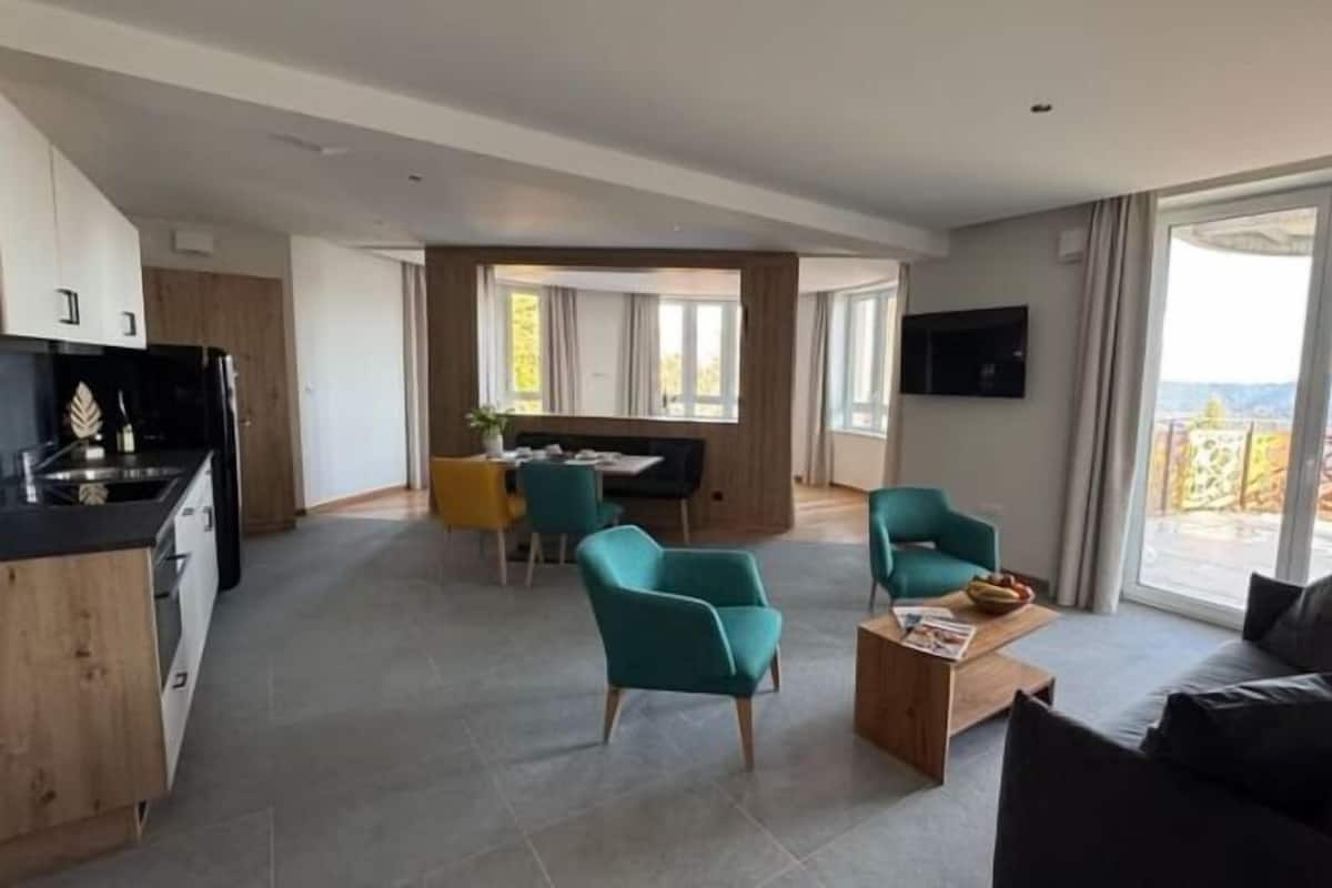 Sala de estar moderna com sofá, poltronas verde-água e mesa de centro. Há uma área de jantar com cadeiras amarelas e uma mesa. Cozinha com bancada e armários pretos é visível. Grandes janelas oferecem vista do exterior.