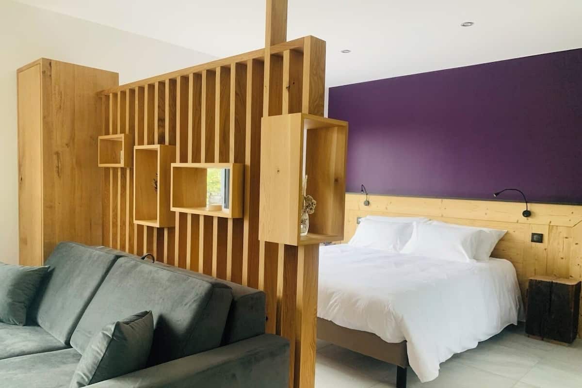 Quarto de hotel moderno com divisória de madeira, sofá cinza, cama com roupa de cama branca e parede de destaque roxa.