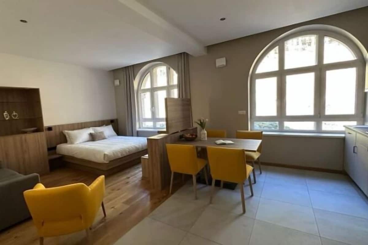 Quarto de hotel moderno com cama, sofá, mesa de jantar com cadeiras e janelas amplas.