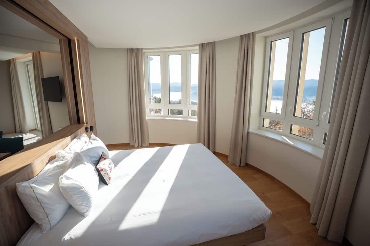 Quarto de hotel com uma cama grande, cabeceira de madeira, televisão de tela plana e vista para as montanhas pelas janelas.