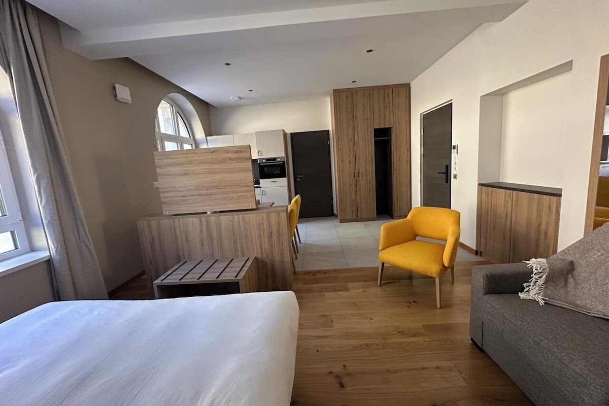 Quarto de hotel moderno com uma cama grande, um sofá, uma mesa de centro de madeira e uma poltrona amarela. Há uma kitchenette ao fundo e um guarda-roupa de madeira.