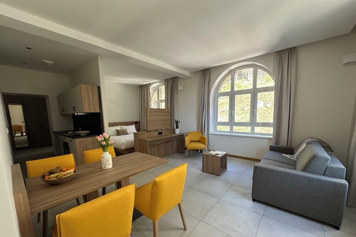 Uma sala de estar moderna com área de jantar, um sofá cinza, cadeiras amarelas e uma cozinha ao fundo.
