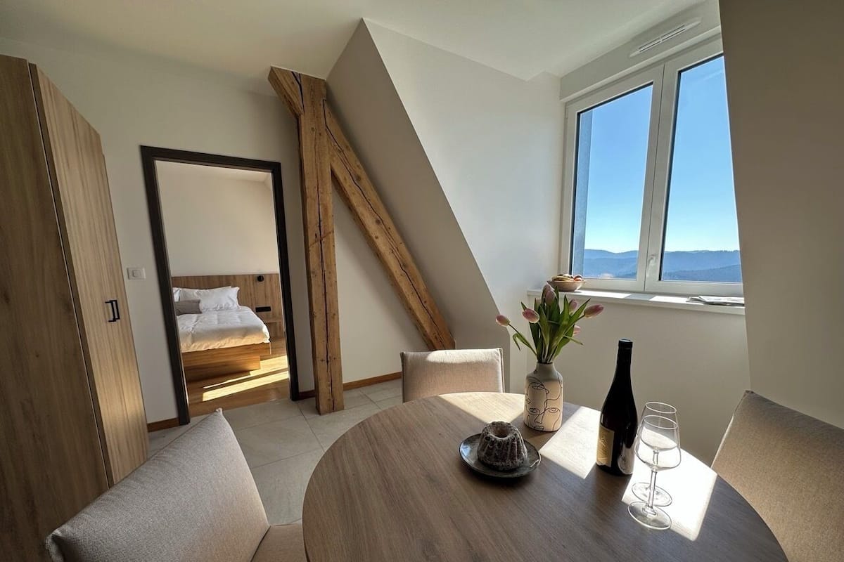 Quarto moderno com cama, mesa de jantar, garrafa e copos, e vista para o exterior.