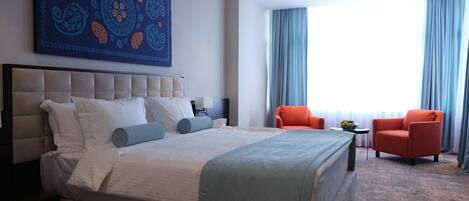 Quarto de hotel moderno com uma cama grande, duas cadeiras, uma escrivaninha e uma janela ampla com cortinas.