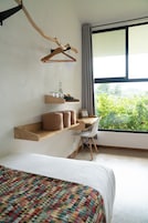 Quarto de hotel com cama, escrivaninha, cadeira, janela com vista para área verde e prateleira de madeira com objetos.