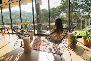 Varanda espaçosa com vista para uma paisagem arborizada, mobiliada com duas cadeiras de arame pretas e um tapete tecido colorido.