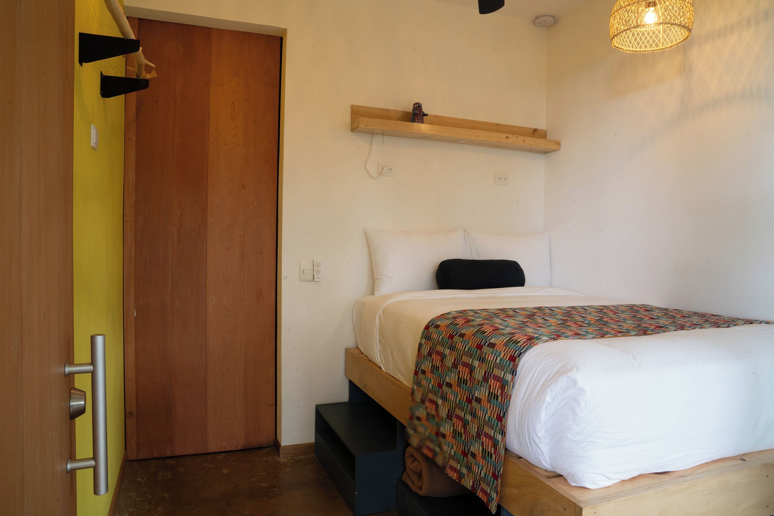 Quarto com cama, cabeceira de madeira, prateleira acima da cama, porta e luminária.