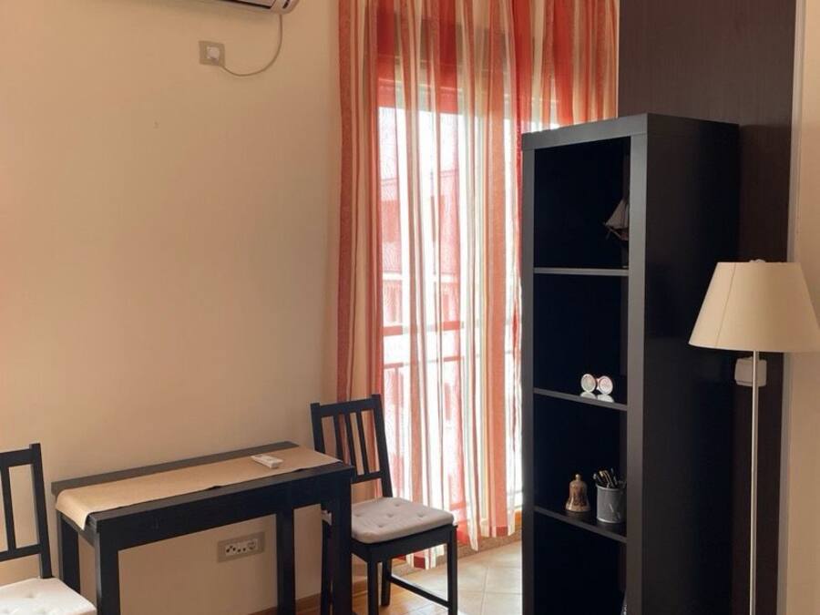 Um quarto com piso de madeira, uma mesa de jantar com cadeiras, uma estante preta e um abajur.