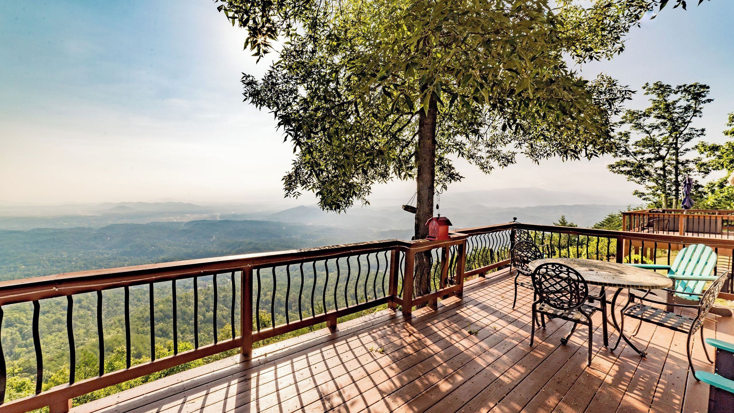 Um deck de madeira com corrimão, uma mesa e cadeiras, com vista para uma paisagem montanhosa.
