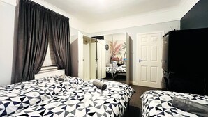 Quarto de hotel com duas camas, uma janela grande com cortinas, um guarda-roupa e uma porta que dá para outro cômodo.