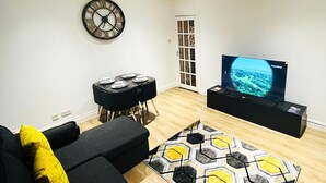 Uma sala de estar com um sofá preto, uma almofada amarela, uma mesa de centro preta, um rack de TV preto com uma TV exibindo um canal de viagens, e um tapete estampado.