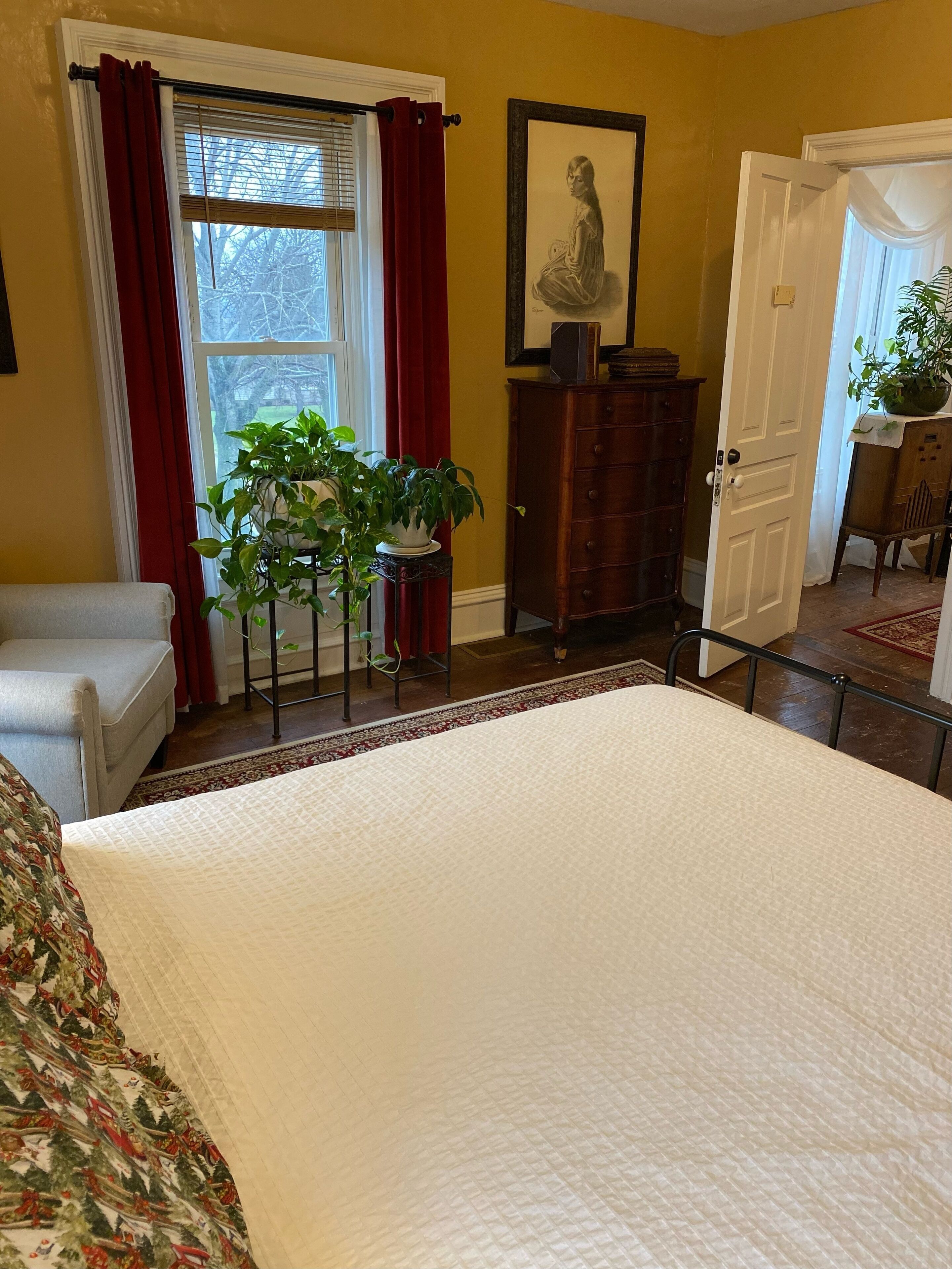 Quarto com uma cama grande, penteadeira, cadeira, janela com cortinas vermelhas e planta em vaso.
