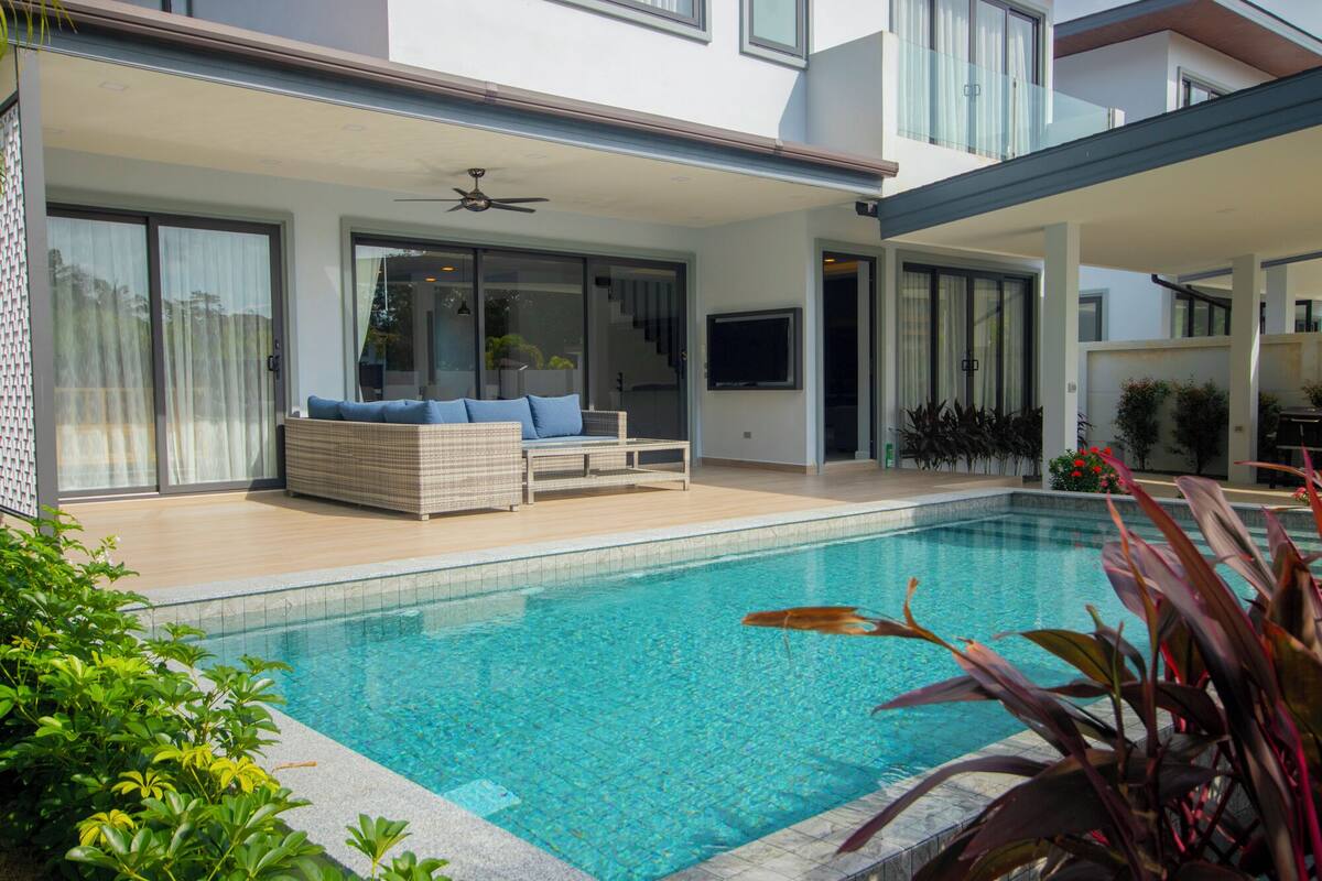 Uma casa moderna com piscina, um alpendre coberto com mobiliário de exterior e uma televisão grande fixada na parede.