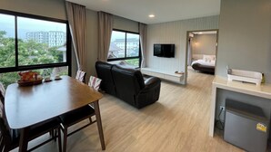 Quarto de hotel moderno com área de refeições, sofá, TV fixada na parede e vista para a paisagem urbana.