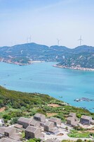 Uma vila costeira com edifícios de pedra tradicionais, mar azul cristalino e turbinas eólicas ao fundo.