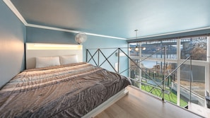 Quarto com cama, cabeceira, janela com vista para uma ponte e vegetação, e luminária pendente.