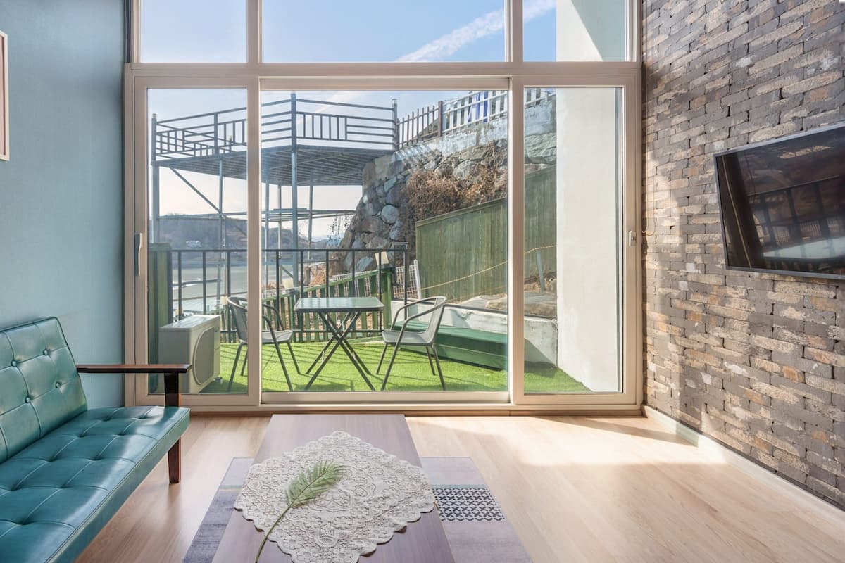 Um quarto com sofá verde, mesa de centro de madeira e uma grande porta de vidro deslizante que dá para área externa com gramado verde, cerca de metal e parede de pedra.