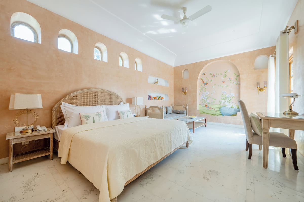 Quarto espaçoso com uma cama grande, uma escrivaninha e uma cadeira. Há várias janelas e um ventilador de teto.
