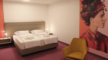 Quarto de hotel com cama, mesa de cabeceira, abajur, poltrona amarela e um grande retrato na parede.