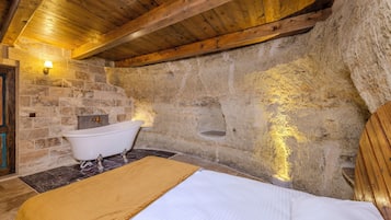 Casa de banho rústica com parede de pedra, teto de madeira, banheira branca e cama com manta bege.