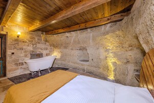 Casa de banho rústica com parede de pedra, teto de madeira, banheira branca e cama com manta bege.