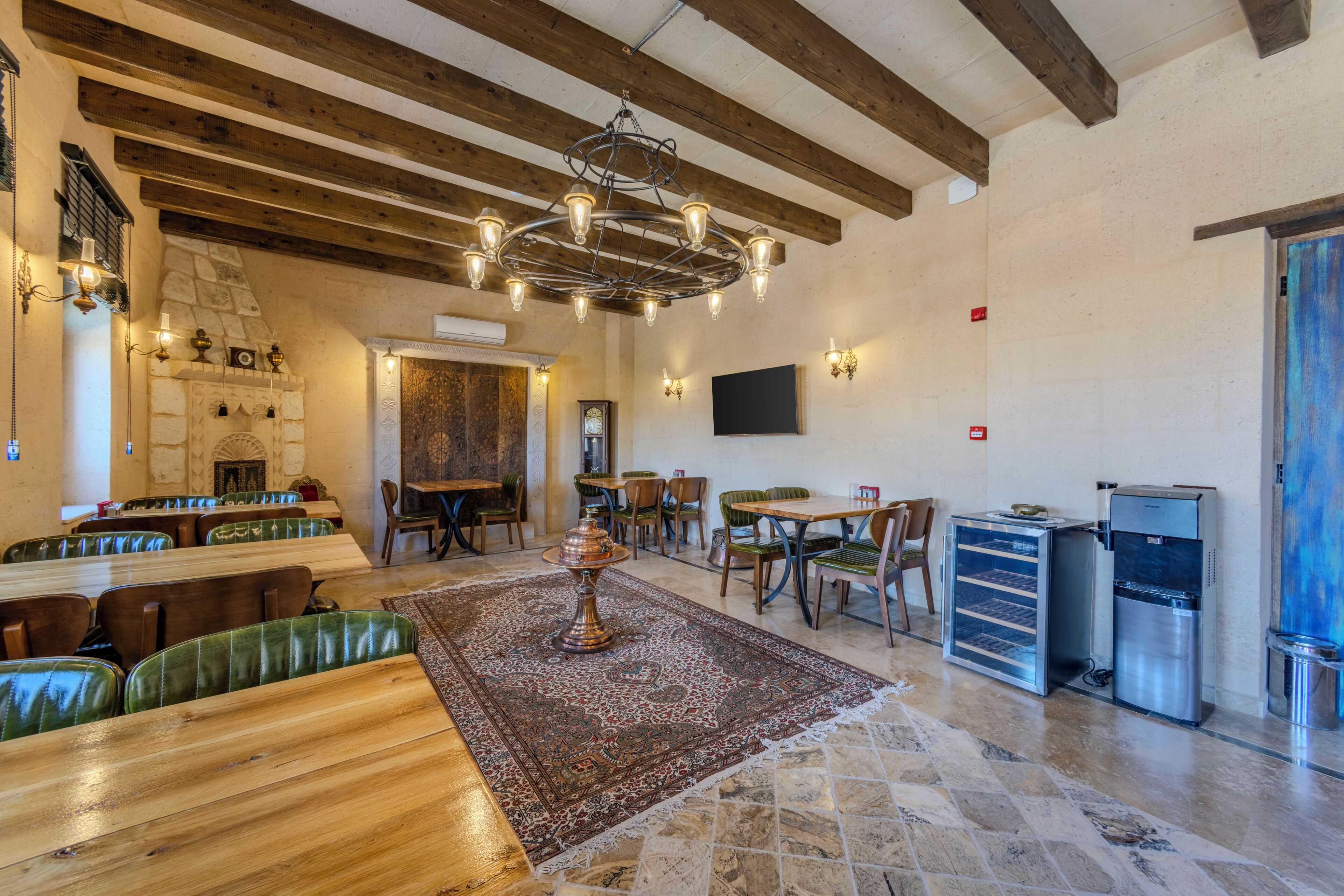 Uma ampla área de refeições com mesas de madeira e cadeiras estofadas verdes, um centro de mesa decorativo em cobre e um tapete grande com padrão.