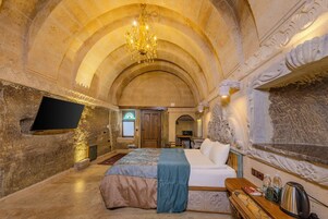 Quarto de hotel com teto abobadado, lustre, cama com colcha azul e bege, mesa de cabeceira de madeira com chaleira e televisão de tela plana na parede.