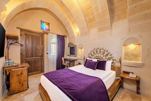 Quarto de hotel com uma cama grande, uma cômoda de madeira, uma mesinha de vaidosa com espelho e uma parede de pedra.
