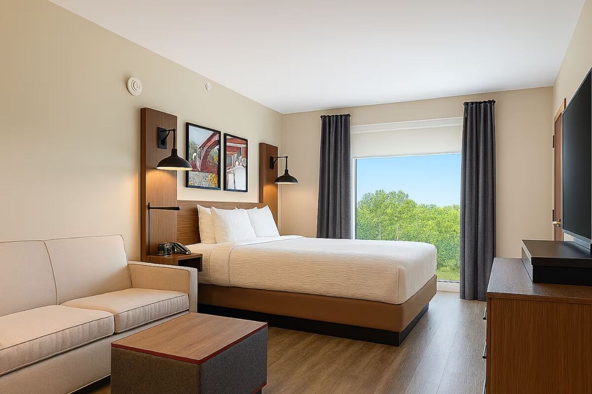 Quarto de hotel com cama, sofá, escrivaninha, televisão e janela com cortinas.