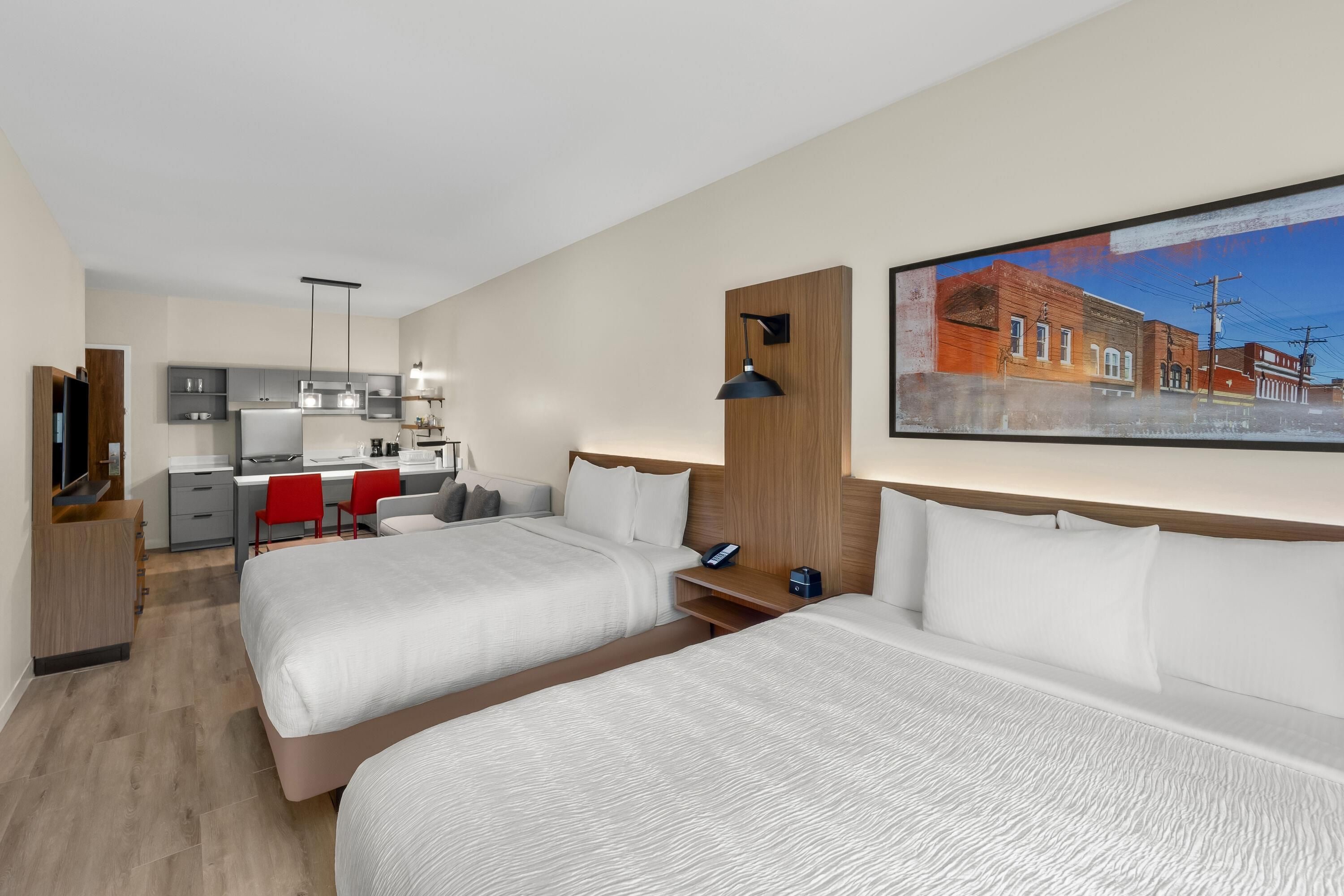 Quarto de hotel com duas camas, uma escrivaninha, uma cadeira, uma televisão e um quadro de uma cena urbana.