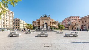 Uma ampla praça pública com um monumento central, rodeada por arquitetura clássica e assentos modernos.