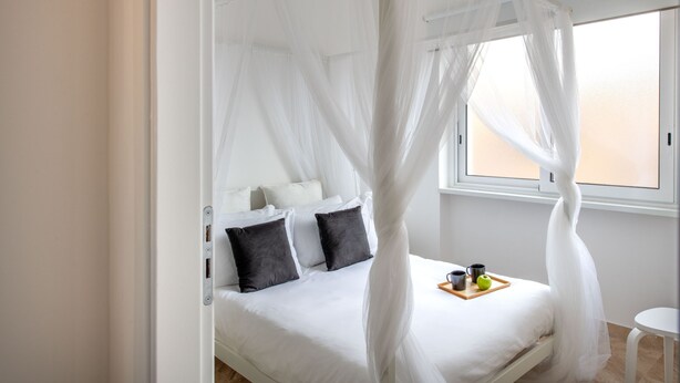 Quarto com cama de dossel, roupa de cama branca e mesa de cabeceira com um serviço de objetos.