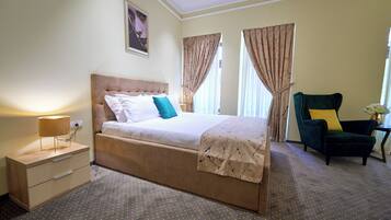 Quarto de hotel com uma cama grande, mesa de cabeceira, uma cadeira e cortinas.