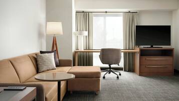 Quarto de hotel com sofá, apoio para os pés, escrivaninha, cadeira, televisão, abajur e uma mesinha.